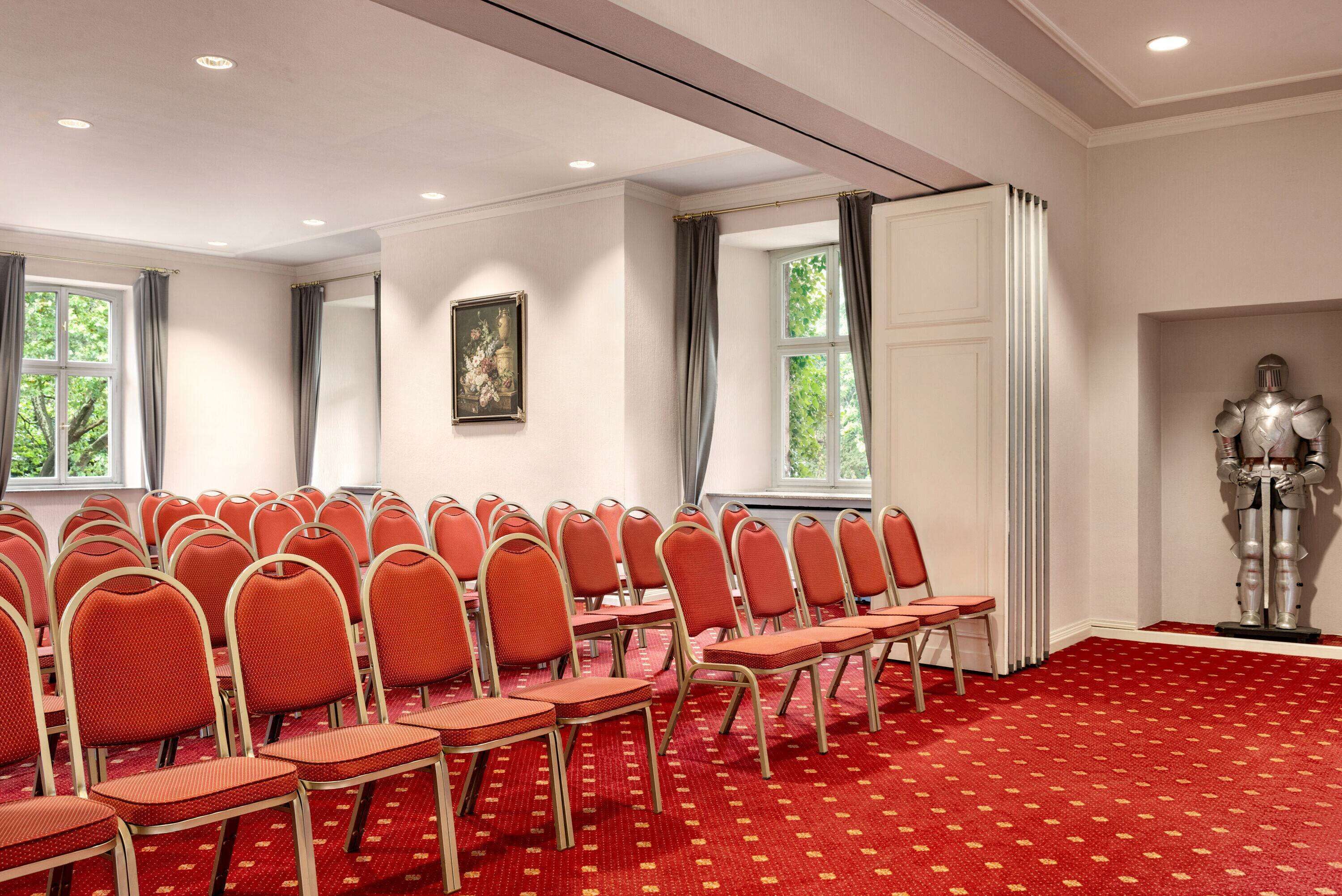 Sala de conferências com fileiras de cadeiras vermelhas, uma estátua e janelas amplas com cortinas.