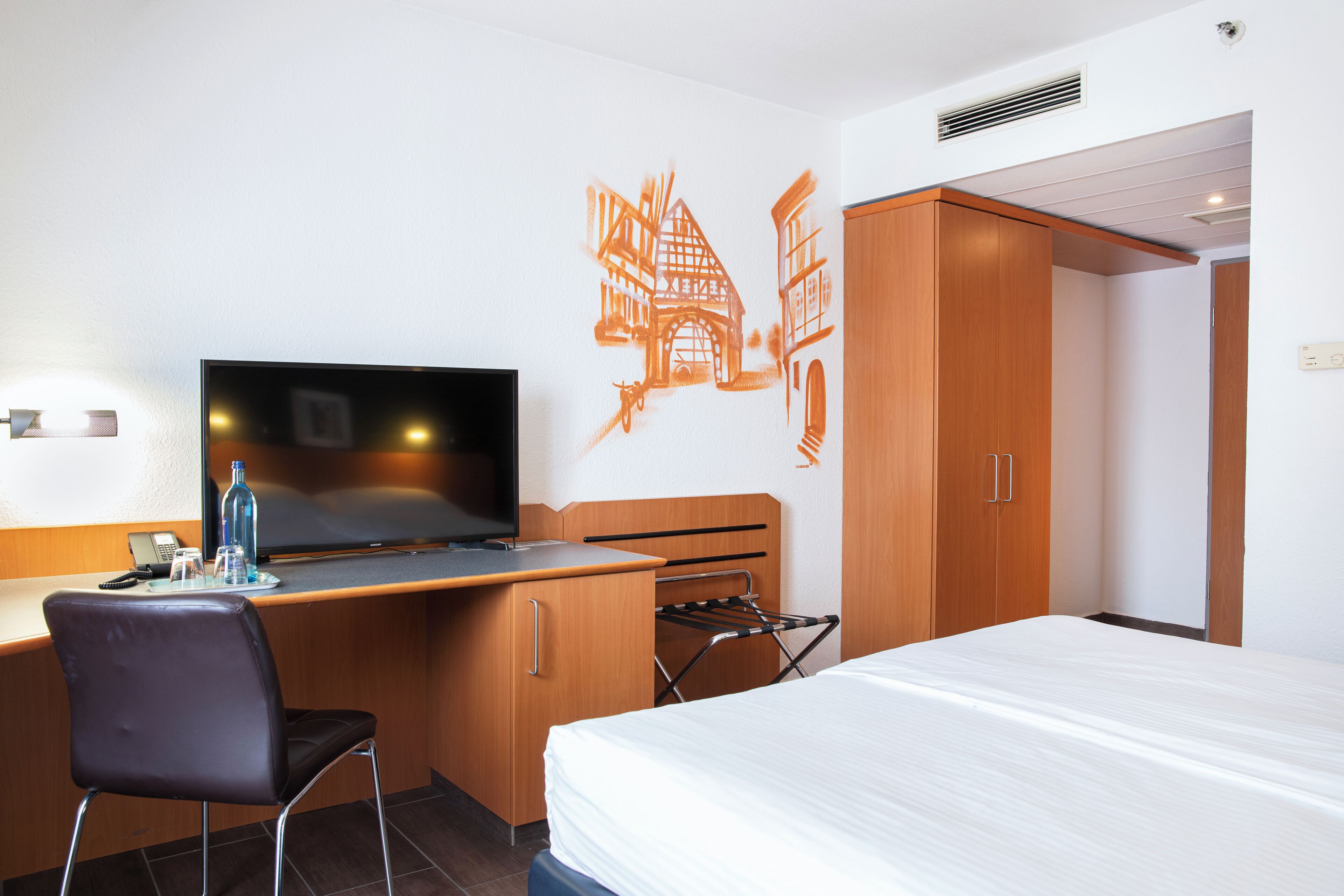 Quarto de hotel com cama, escrivaninha, cadeira, televisão e guarda-roupa.