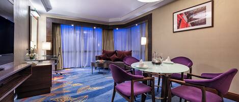 Quarto de hotel com área de refeições, sofá, TV e uma grande janela com cortinas.