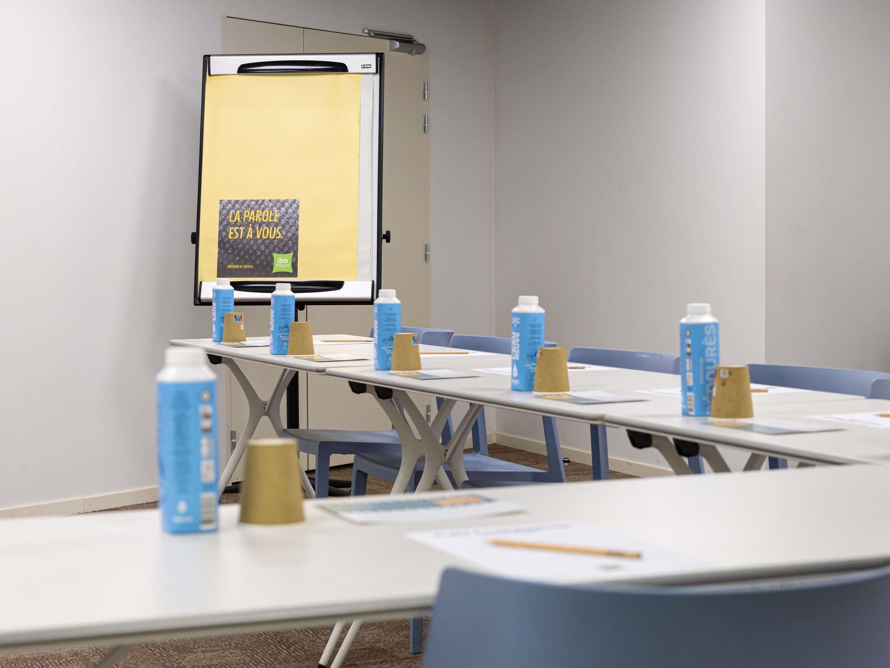 Sala de conferência com mesas, cadeiras, tela de projeção e garrafas de água.