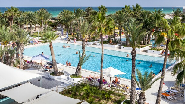 Uma área de piscina em resort, com várias piscinas, rodeada por palmeiras e espreguiçadeiras.