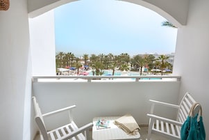 Varanda com cadeiras brancas e uma mesa, com vista para uma área de piscina com palmeiras e guarda-sóis.