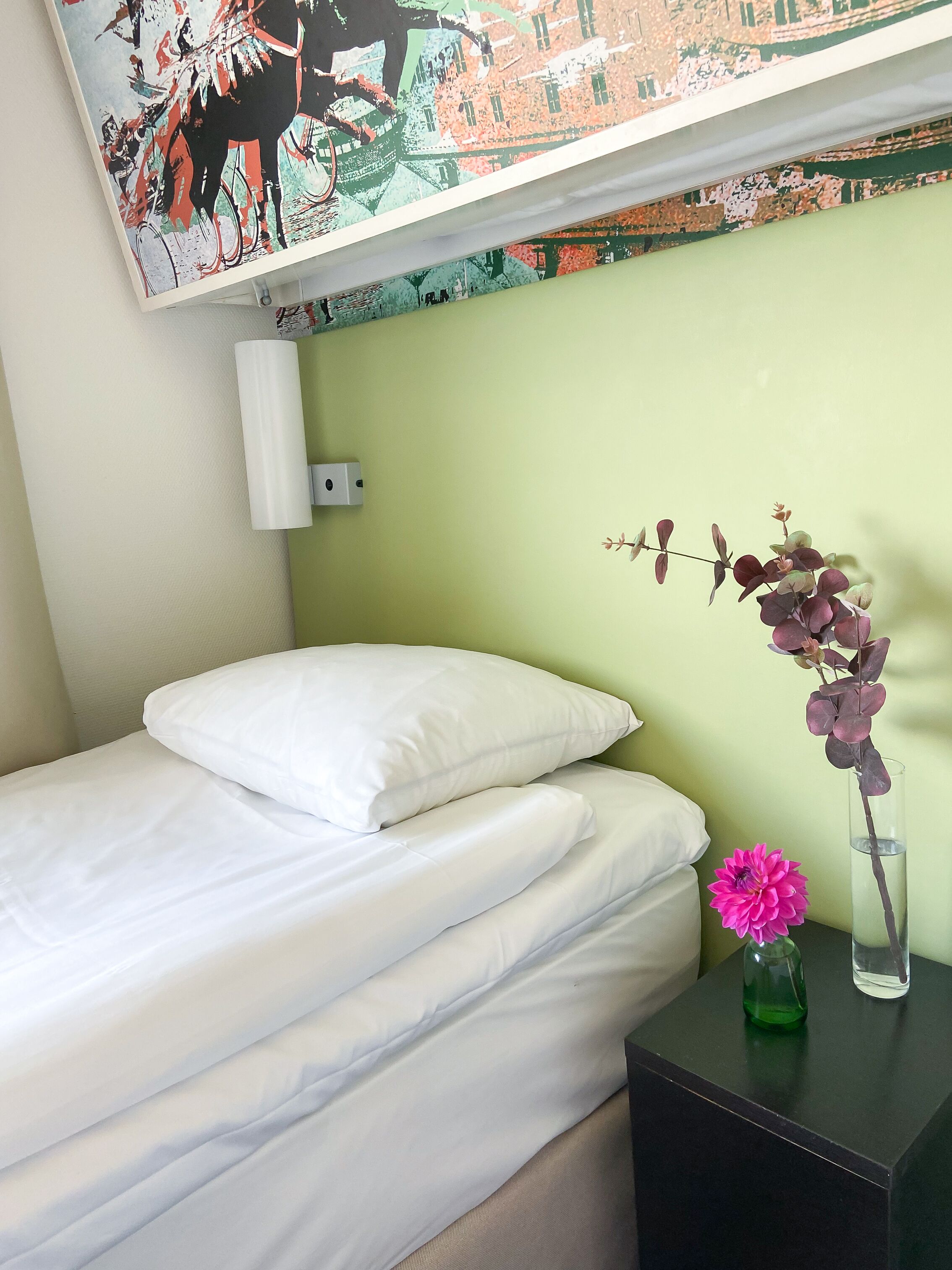 Cama bem arrumada com roupa de cama branca, parede verde, uma pequena mesa com um vaso de flores e um luminário fixado na parede.