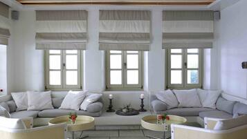 Uma sala de estar moderna com um sofá seccional, duas mesas laterais e janelas grandes com cortinas brancas.