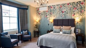Quarto com uma cama grande, duas poltronas, um candelabro, papel de parede floral e uma janela com cortinas.