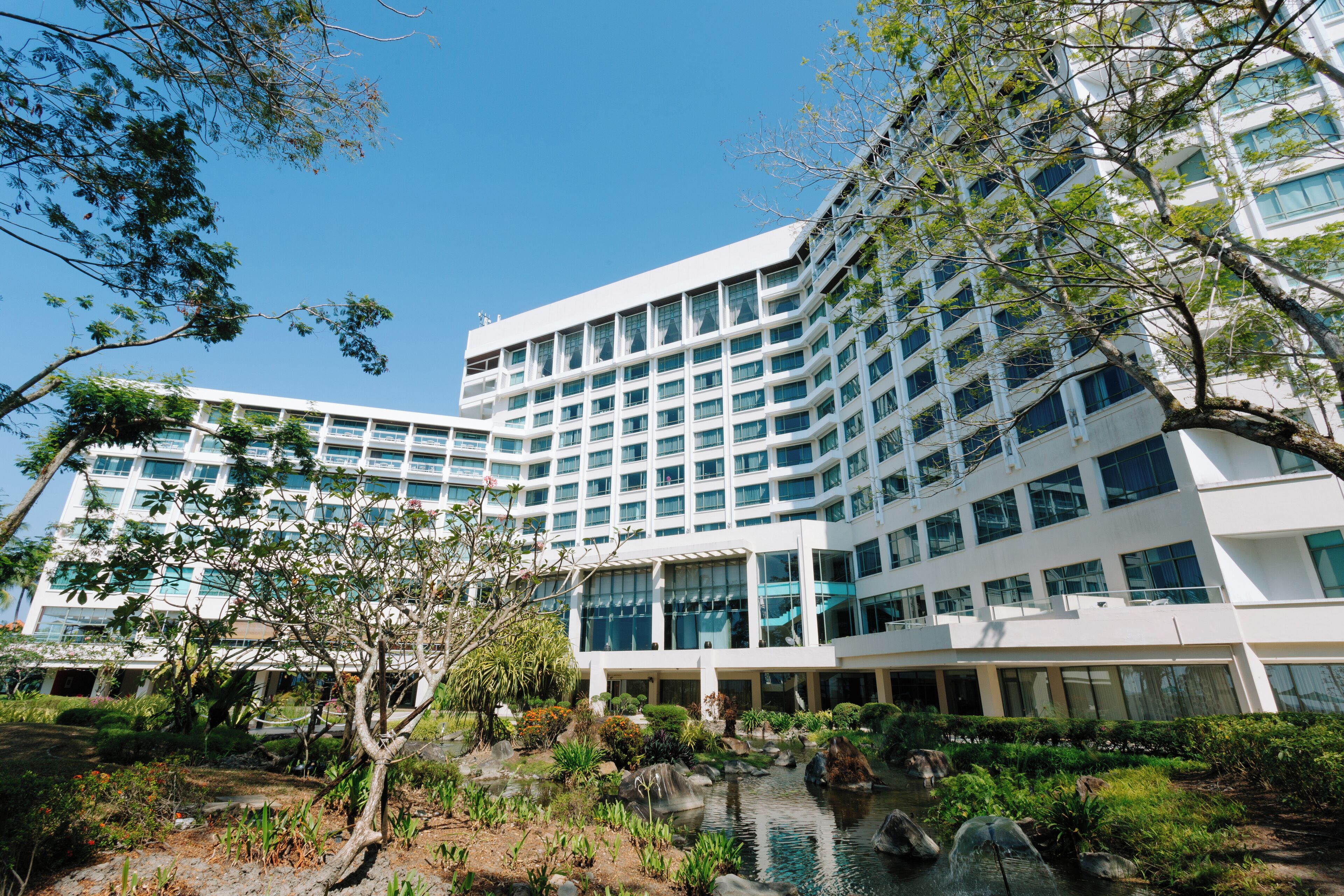 Um hotel moderno com vários andares, grandes janelas e um jardim paisagístico com lago.
