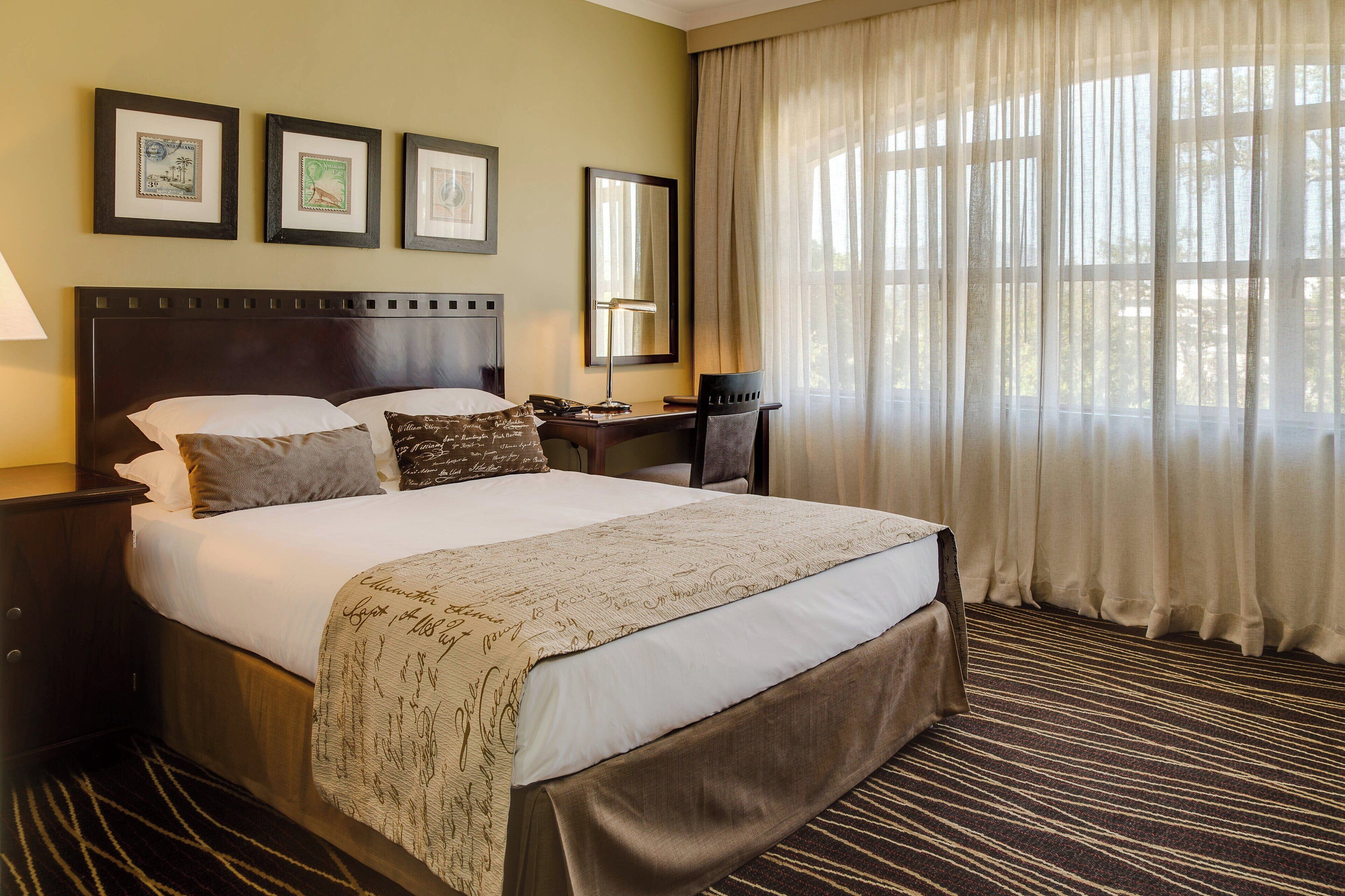 Quarto de hotel com uma cama grande, uma escrivaninha, uma cadeira, uma mesinha de cabeceira e uma janela com cortinas.