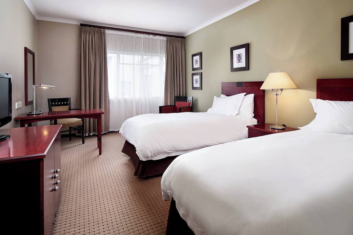 Quarto de hotel com duas camas, uma escrivaninha, uma cadeira, uma televisão e uma luminária.