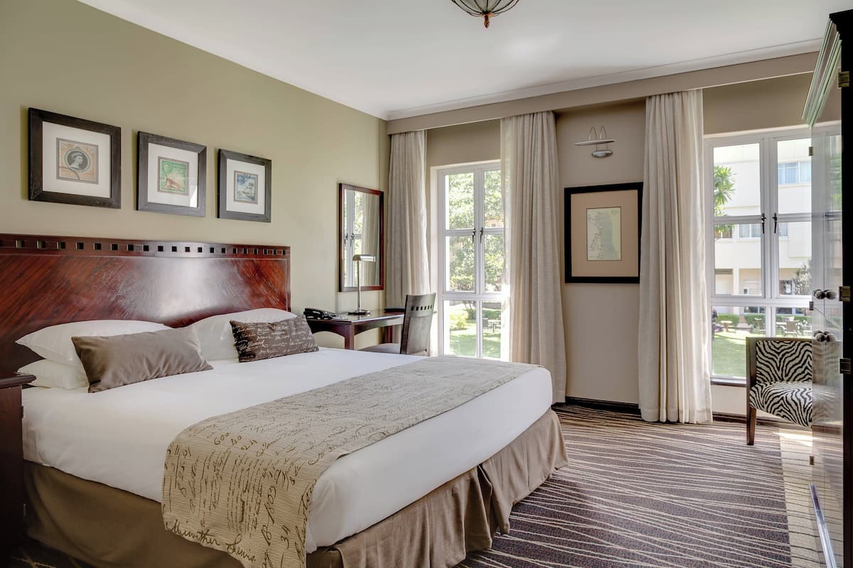 Quarto de hotel com cama grande, escrivaninha e cadeira. Três quadros emoldurados na parede, janela com cortinas e vista para o exterior.