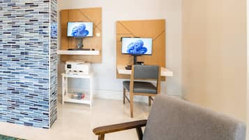 Um espaço de escritório moderno com duas mesas, cada uma com um monitor de computador e teclado, e uma cadeira de escritório cinzenta. Há uma estante branca com uma impressora e um livro. A parede apresenta um design de azulejos com padrão azul e branco.