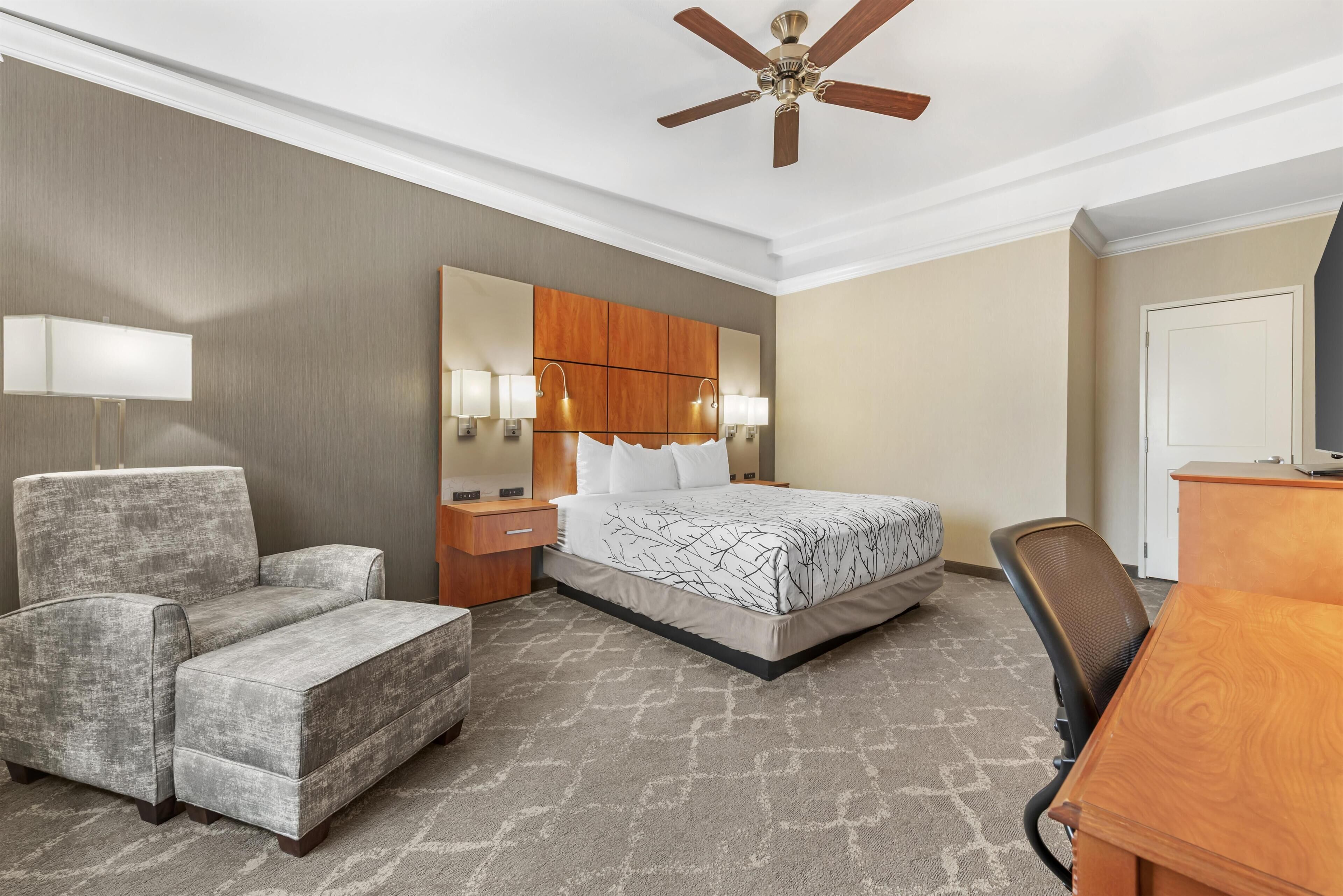 Quarto de hotel com cama, escrivaninha, cadeira, ventoinha no teto e mesa de cabeceira com luminárias.