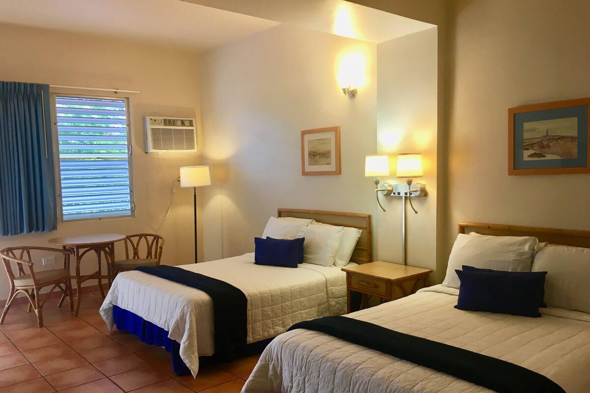 Quarto de hotel com duas camas, uma escrivaninha, uma cadeira e uma janela com persianas.
