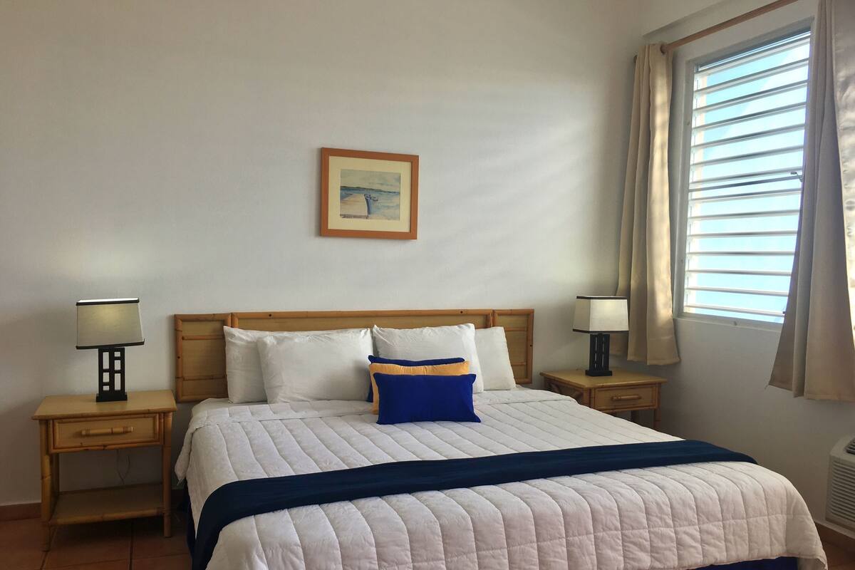 Uma cama bem arrumada com um edredom azul e branco, duas mesas de cabeceira com abajures, um quadro na parede e uma janela com cortinas.