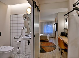 Casa de banho de hotel com sanita, lavatório e chuveiro. No quarto ao lado, há uma cama e uma secretária com uma cadeira.