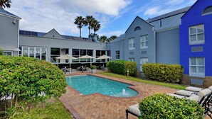 Uma área de piscina com um terraço de tijolos, espreguiçadeiras e um edifício com grandes janelas e painéis solares.