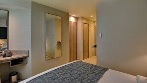Quarto de hotel com uma cama, mesa de cabeceira com telefone, lixeira e espelho. Há portas de madeira e um interruptor de luz na parede.