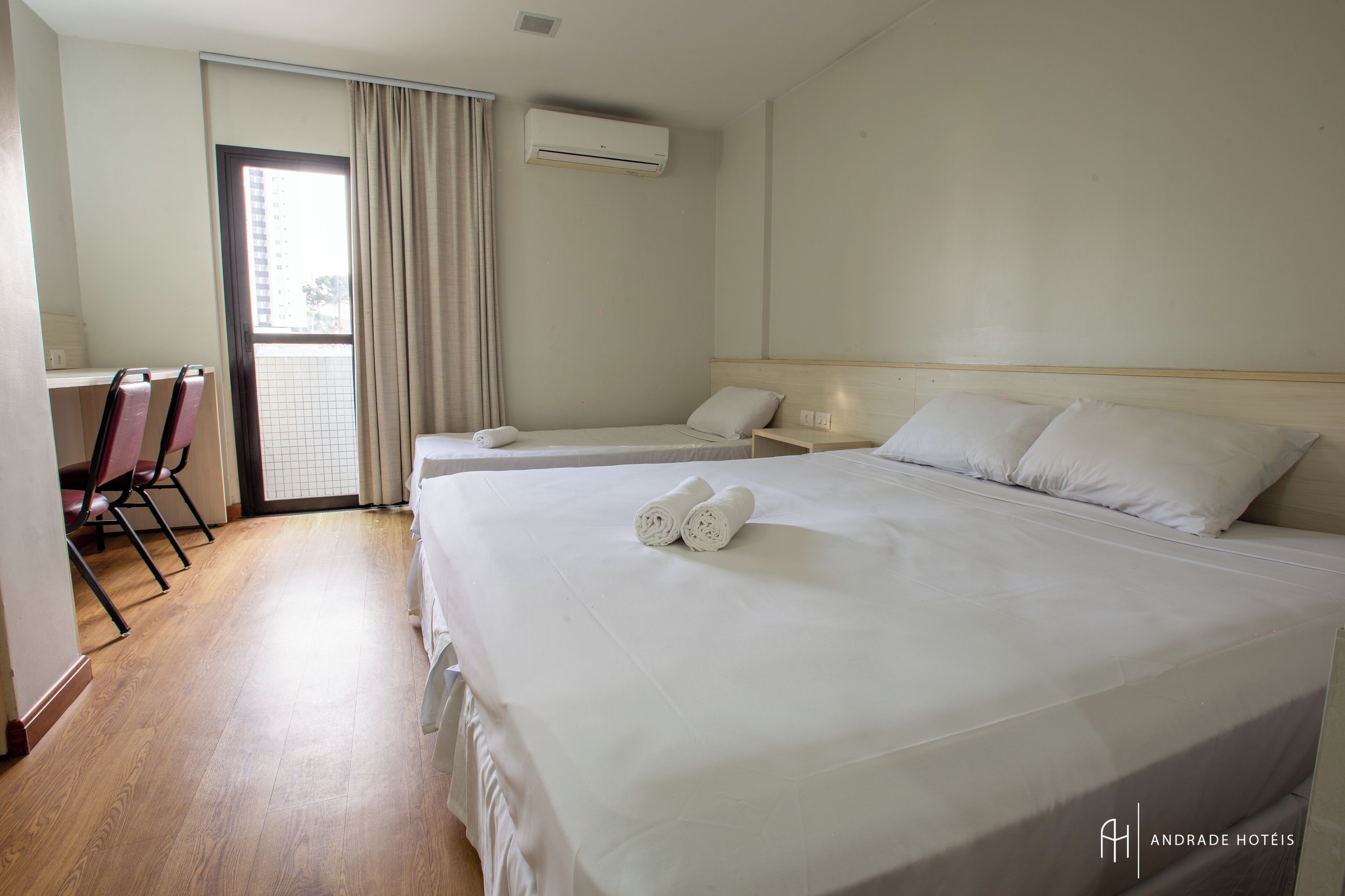 Quarto de hotel com uma cama grande, duas mesas de cabeceira, uma escrivaninha com cadeiras e um ar condicionado.
