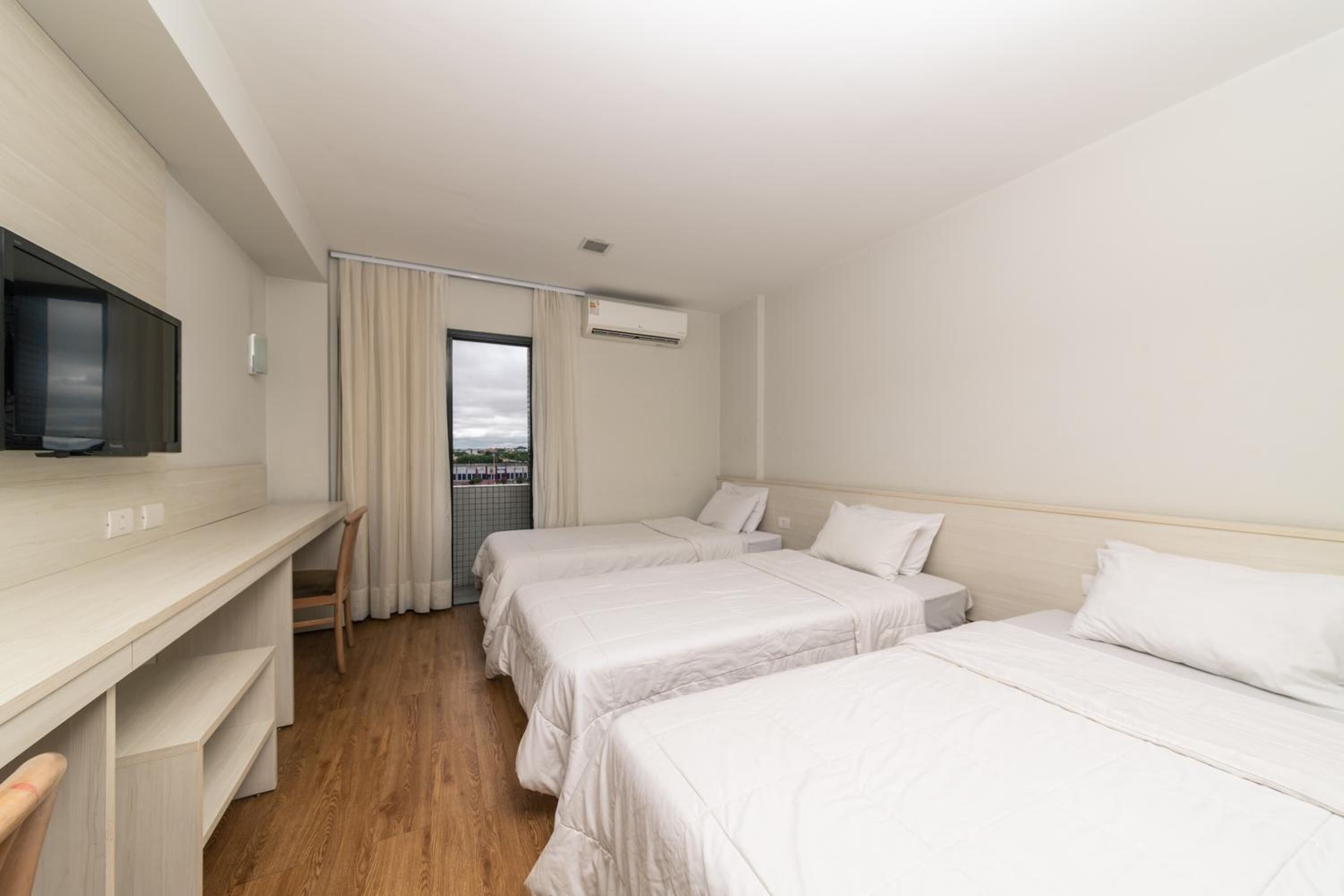 Quarto de hotel com três camas de solteiro, uma escrivaninha com cadeira, televisão e ar condicionado.