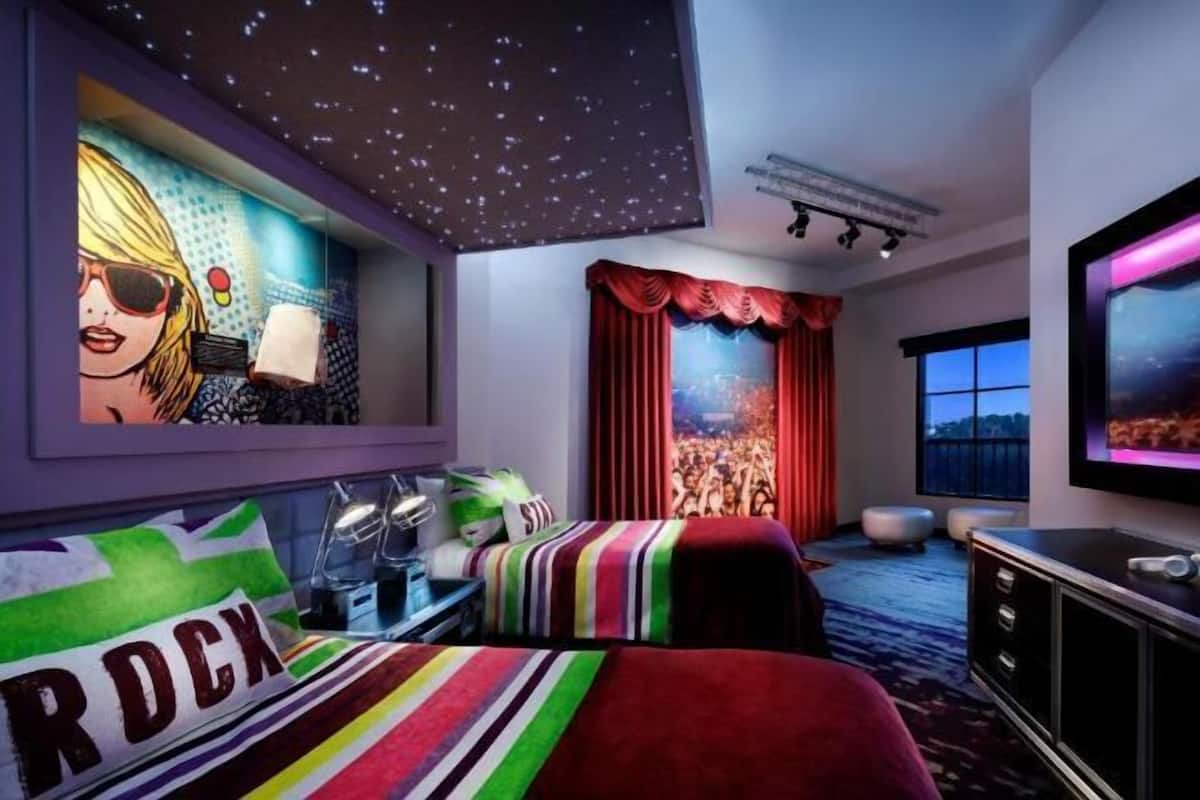 Quarto de hotel com cama, televisão, projetor de teto e janela com cortinas.