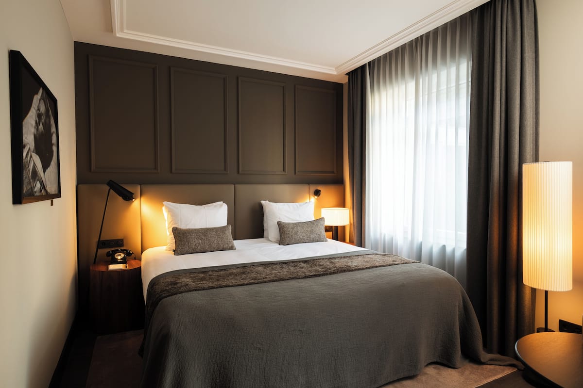 Quarto de hotel com uma cama grande, duas luminárias de cabeceira, um criado-mudo e vista para o exterior através de cortinas translúcidas.