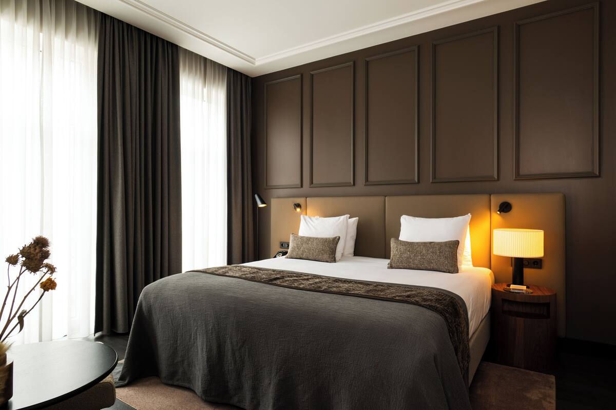 Quarto de hotel com uma cama grande, mesa de cabeceira, abajur e vista para o exterior através de cortinas translúcidas.