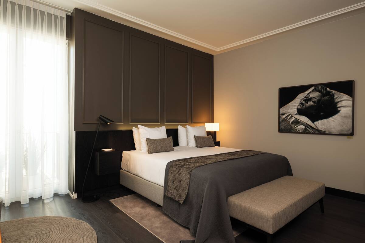 Quarto de hotel moderno com uma cama grande, abajures, um banco e uma pintura na parede.