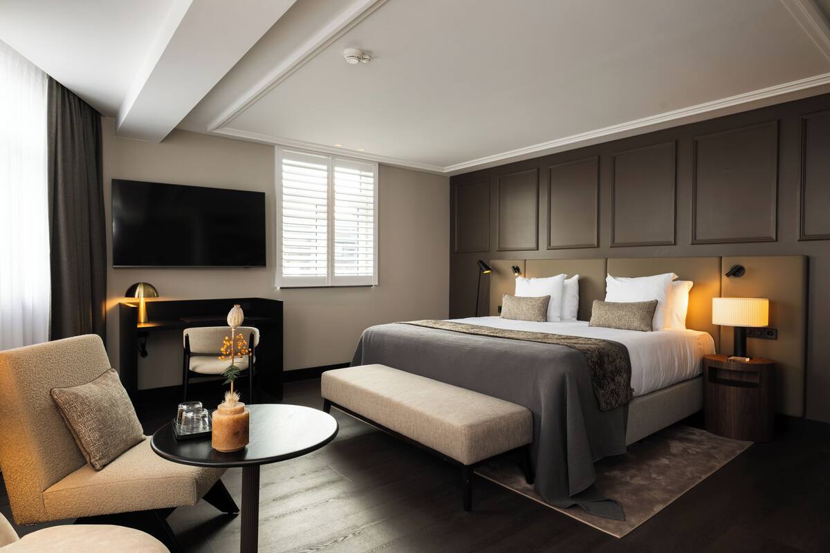 Quarto de hotel moderno com uma cama grande, área de estar com um banco, televisão de tela plana e escrivaninha com luminária.