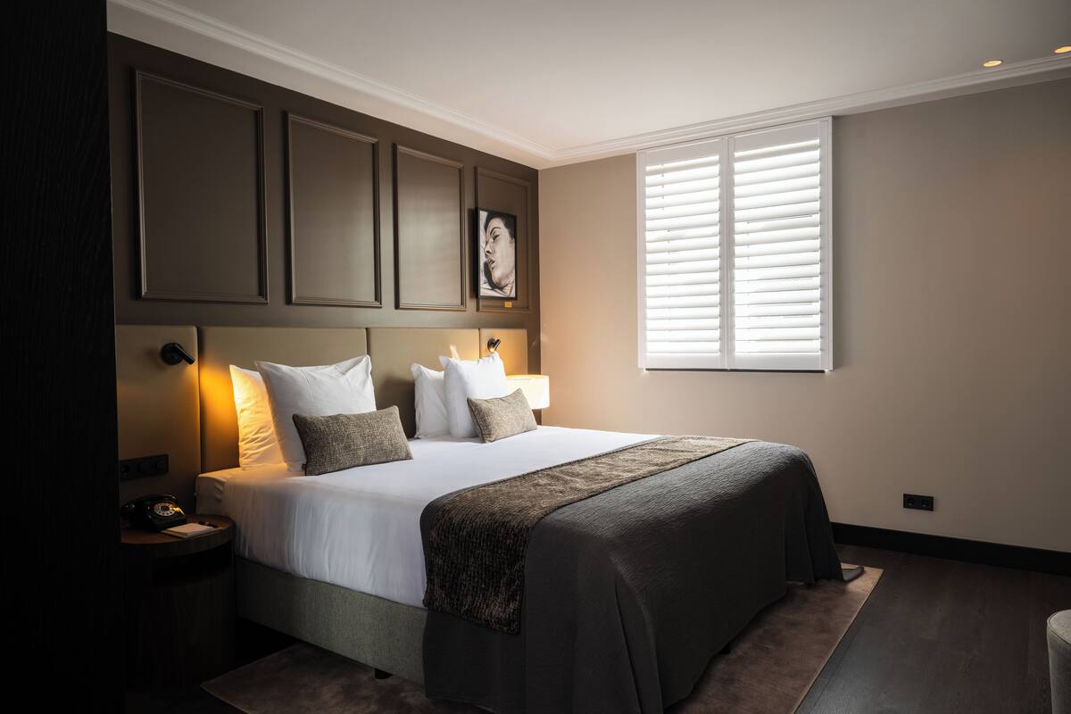 Quarto de hotel bem arrumado com uma cama grande, mesa de cabeceira com abajur e janela com persianas.