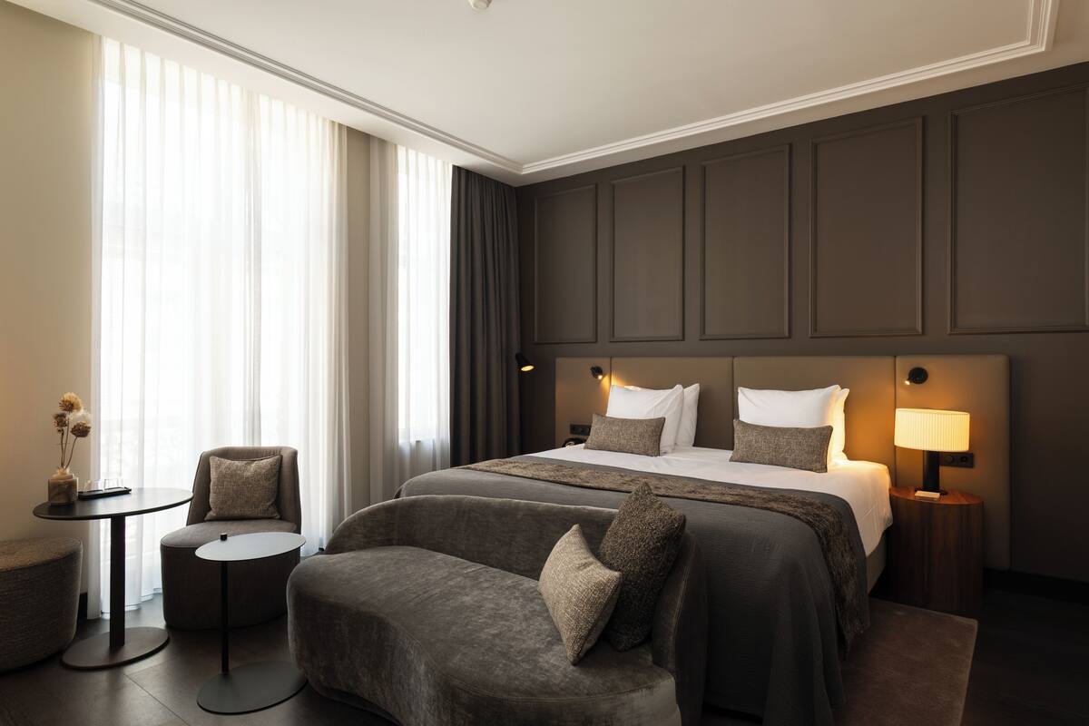 Quarto de hotel com uma cama grande, duas poltronas, uma mesa pequena e uma mesa de cabeceira com abajur.