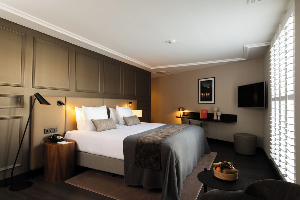 Quarto de hotel moderno com uma cama grande, abajures, televisão de tela plana, uma mesinha redonda com uma fruteira e uma janela com persianas.