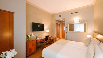 Quarto de hotel com cama, escrivaninha, cadeira, televisão e espelho.