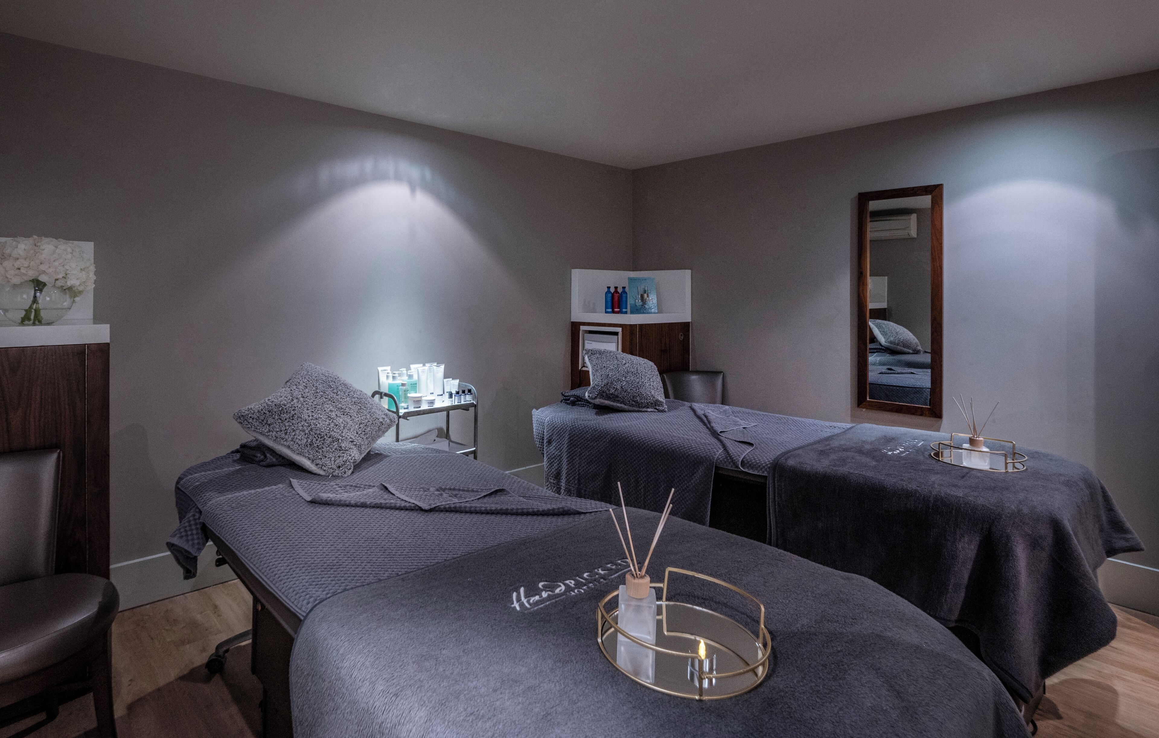 Sala de spa com duas mesas de massagem, uma pequena mesa de escritório com espelho e uma estante com frascos.