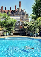 Piscina com uma pessoa nadando, cercada por vegetação exuberante e um edifício histórico com várias chaminés.