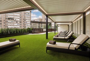 Área de lounge no terraço com grama sintética, espreguiçadeiras e vista para edifícios ao redor.