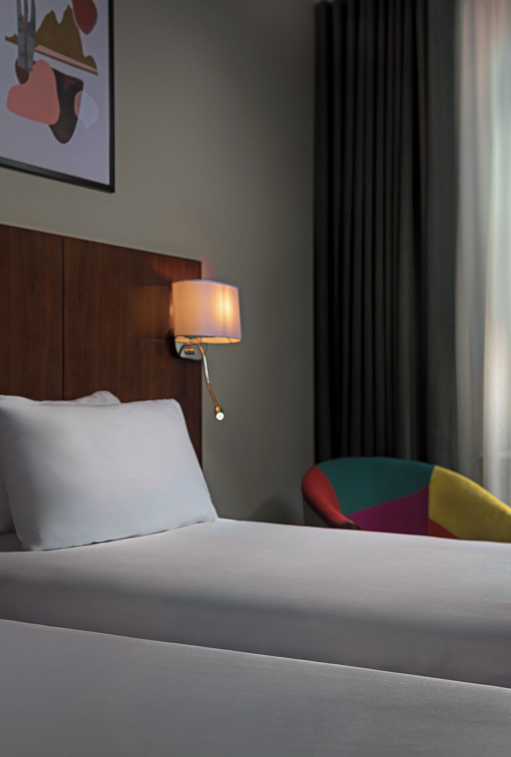 Quarto de hotel com cama, luminária fixada na parede, cadeira colorida e cortina.