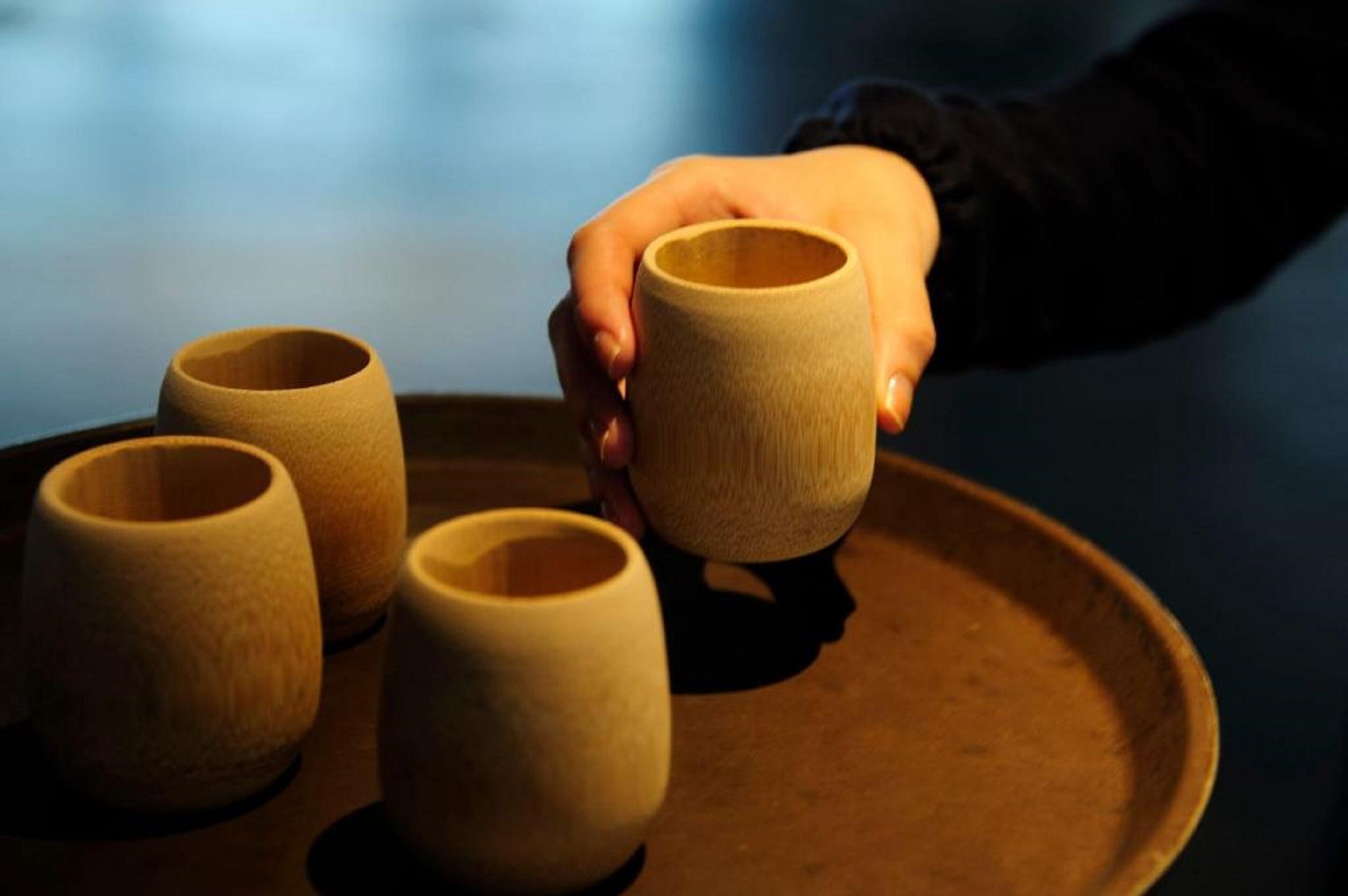 Uma mão segurando uma caneca de cerâmica em um tabuleiro com outras três canecas semelhantes.