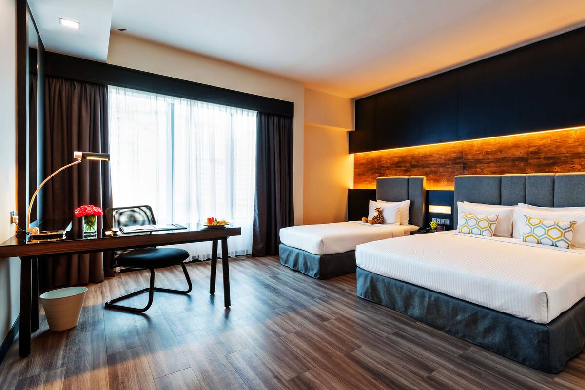 Quarto de hotel com uma cama grande, uma cama menor, uma mesa com luminária e vista para o exterior através de grandes janelas.