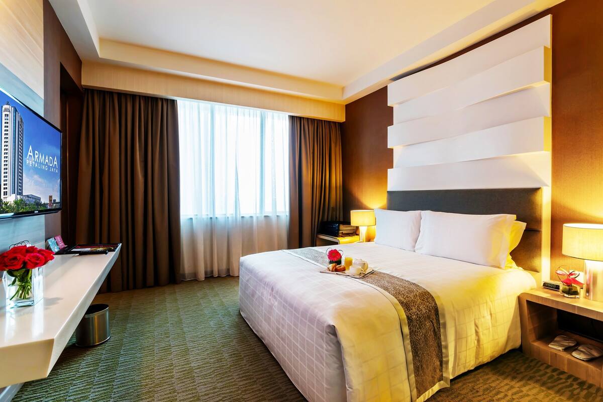Quarto de hotel com uma cama grande, uma escrivaninha com televisão, uma janela com cortinas e uma mesa de cabeceira com luminária.