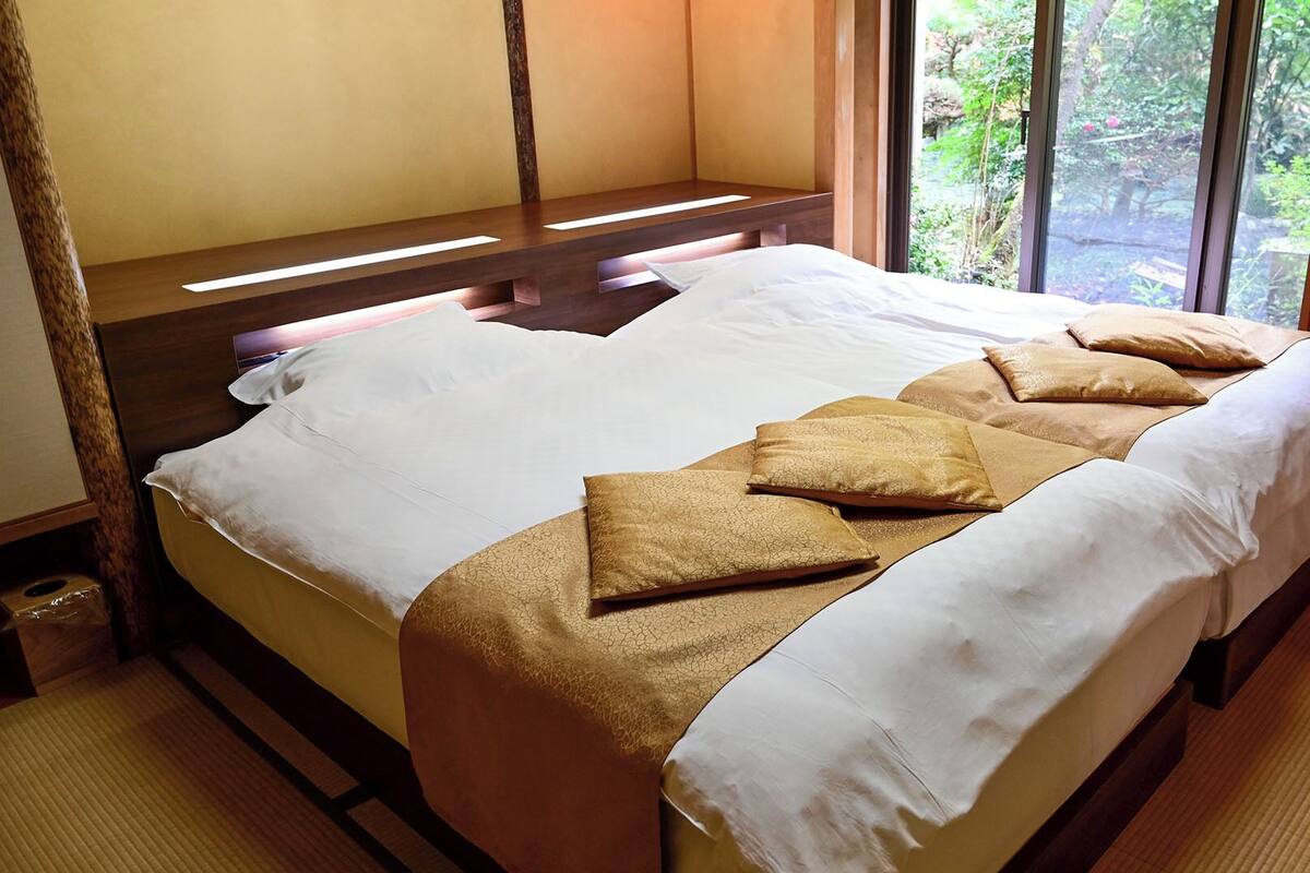 Um quarto em estilo japonês tradicional, com piso de tatame, cama de madeira baixa com roupa de cama branca e vista para o verde através de um painel shoji.