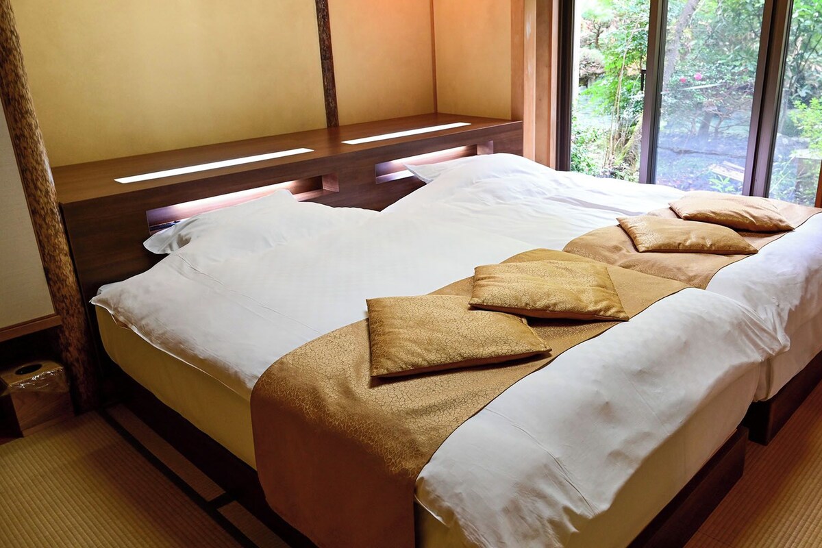Um quarto em estilo japonês tradicional, com piso de tatame, cama de madeira baixa com roupa de cama branca e vista para o verde através de um painel shoji.