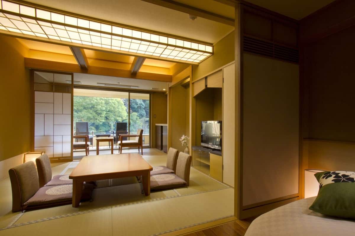 Um quarto japonês tradicional com tatames, uma mesa baixa com almofadas, uma cama com travesseiro e uma televisão.