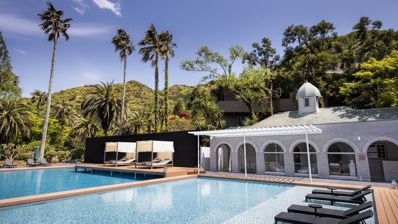 Piscina com espreguiçadeiras, área de estar coberta e um edifício com janelas em arco.
