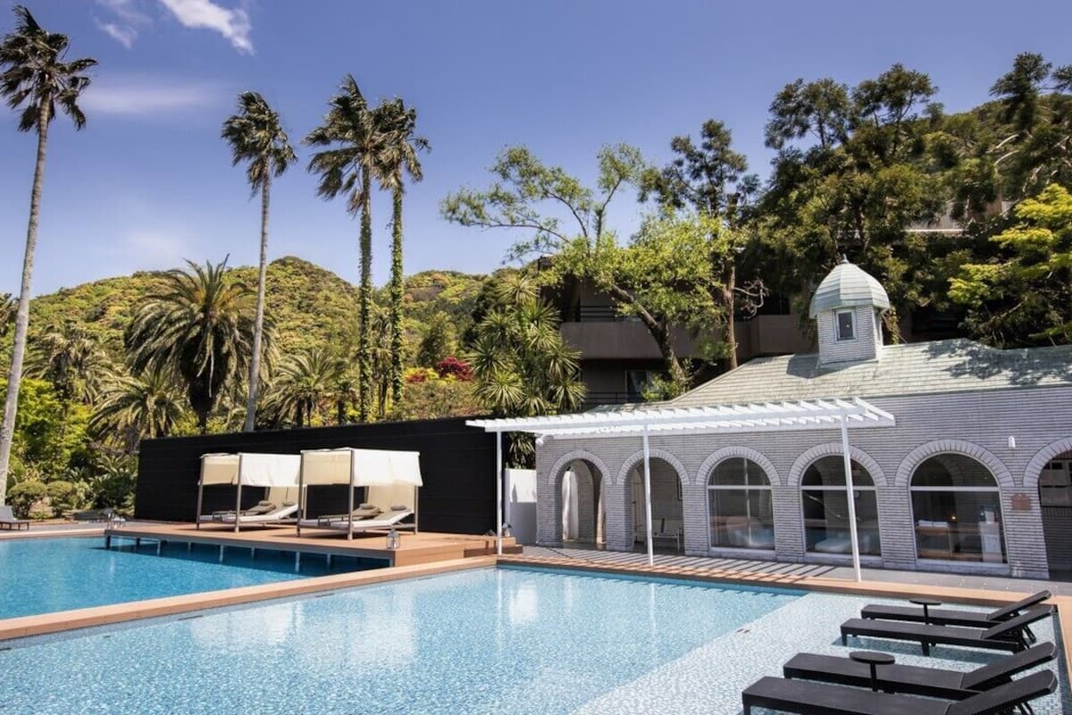 Piscina com espreguiçadeiras, área de estar coberta e um edifício com janelas em arco.