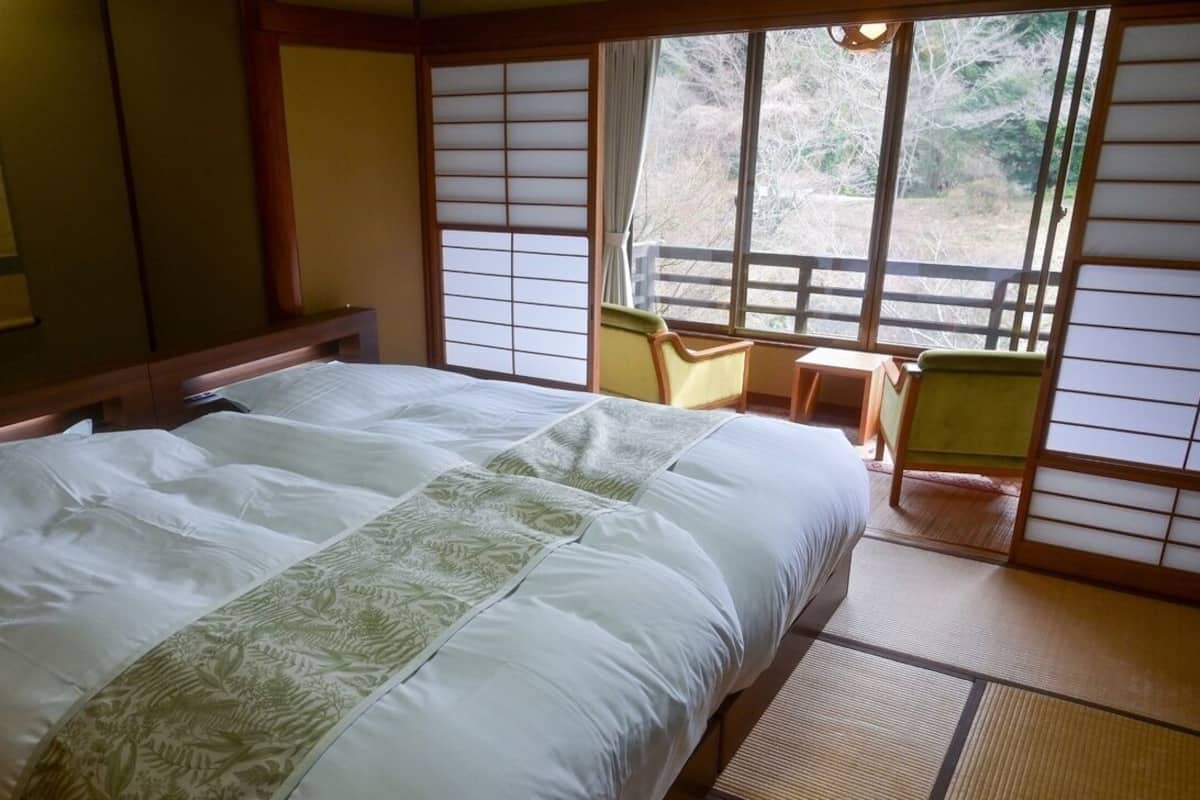Quarto tradicional japonês com piso de tatame, uma cama grande com colcha estampada em verde, uma mesinha e portas corrediças.