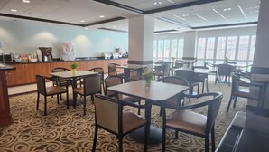Um espaço de refeições de hotel com mesas e cadeiras, um balcão com uma máquina de café e vista para o exterior através de grandes janelas.