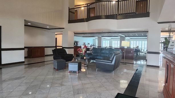 Um hall amplo com teto alto, área de estar com sofás e cadeiras, balcão de recepção e escadaria para um andar superior.
