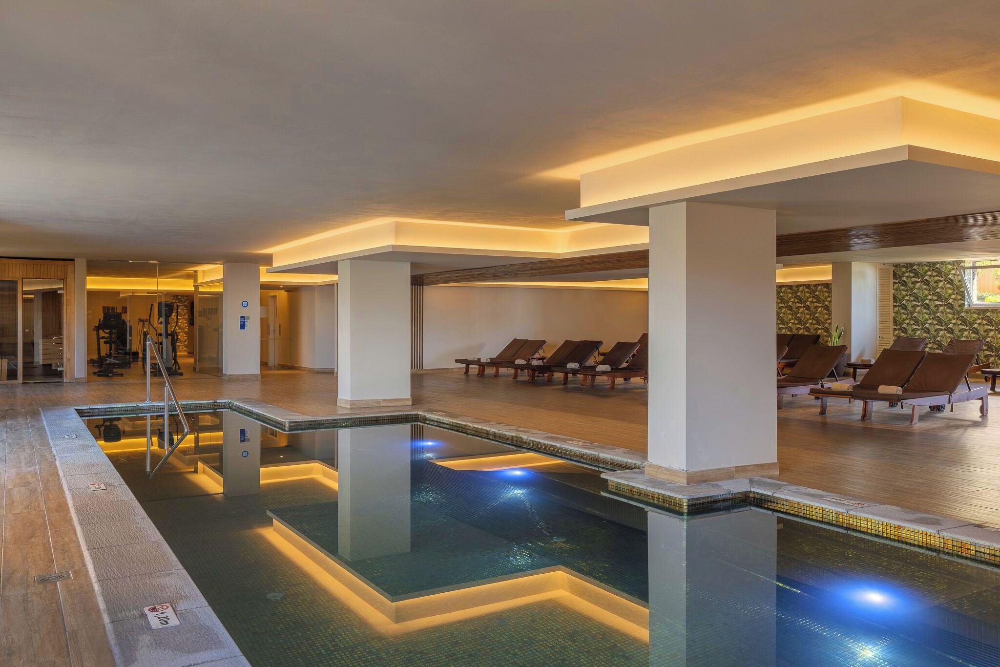 Área interna de piscina moderna com água cristalina, espreguiçadeiras e teto bem iluminado.