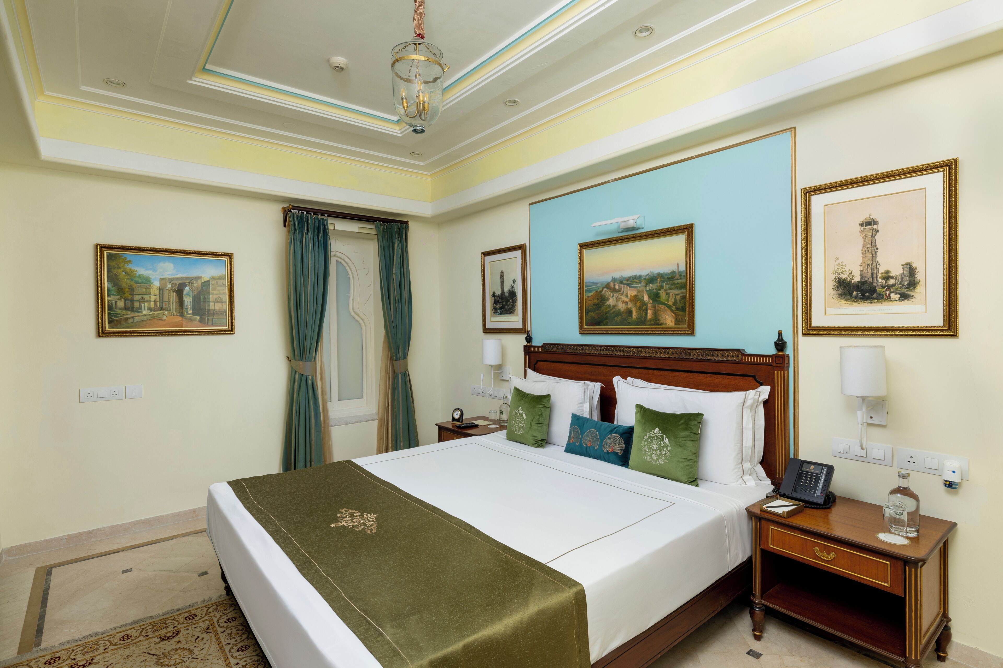 Quarto de hotel com uma cama grande, mesa de cabeceira com luminária, criado-mudo e quadros nas paredes.