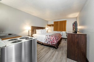 Quarto de hotel com cama, mesa de cabeceira, luminária, TV e cozinha compacta.