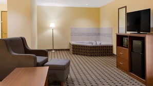 Quarto de hotel com sofá, banquinho, TV de tela plana, geladeira, micro-ondas, pia e banheira.